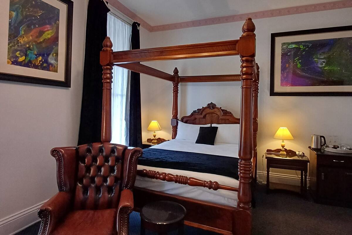 Uma cama de dossel com estrutura de madeira e um candelabro acima. Há uma cadeira com encosto acolchoado e uma pequena mesa redonda ao lado da cama. Duas luminárias estão em mesas de cabeceira, e obras de arte estão expostas nas paredes.