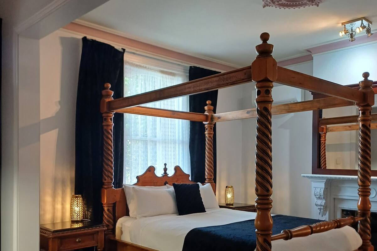 Uma cama com dossel de madeira e design entalhado, uma mesinha de cabeceira com abajur, e uma vista de lareira ao fundo.