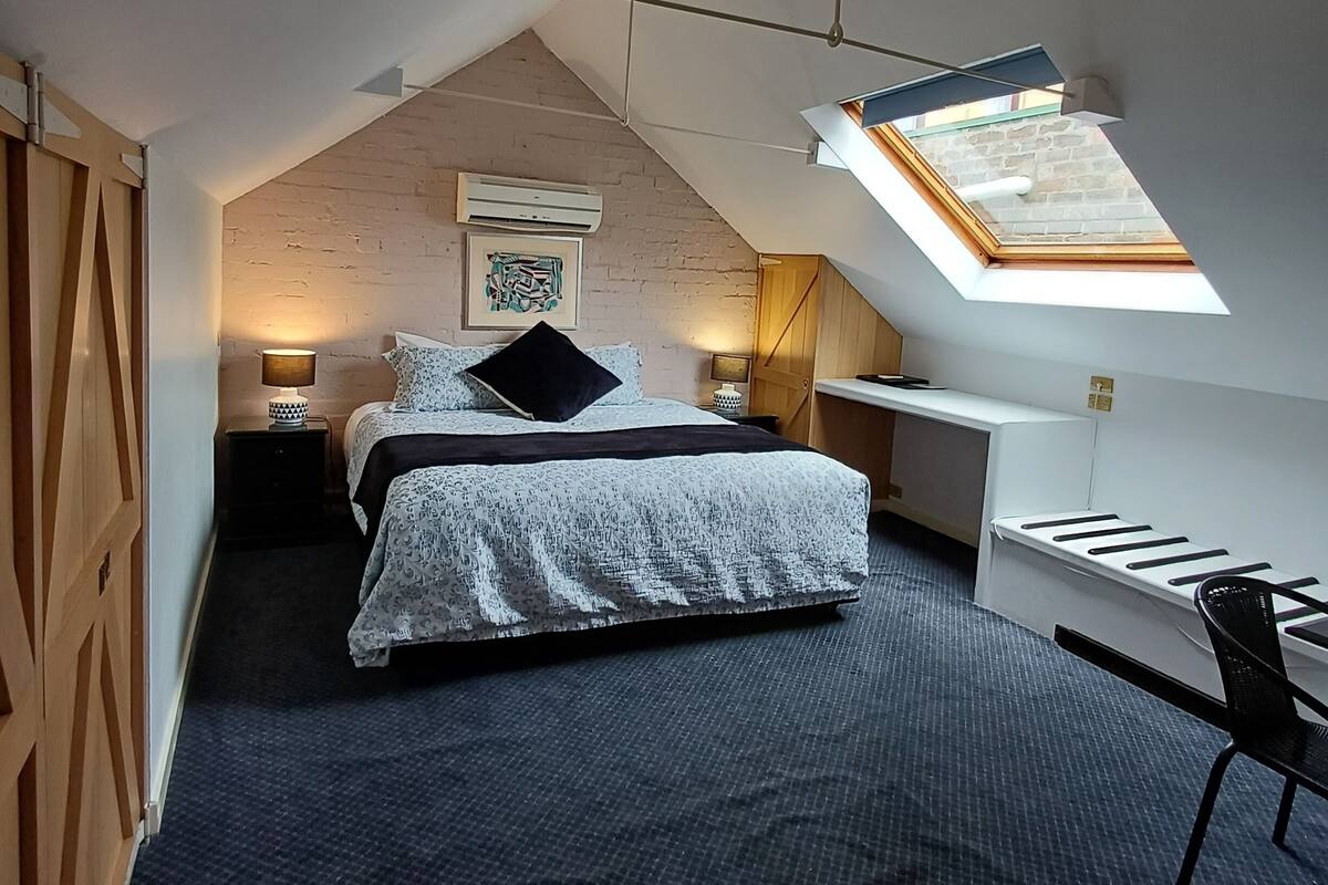 Quarto com teto inclinado, claraboia, cama com roupa de cama estampada, escrivaninha e uma cadeira.