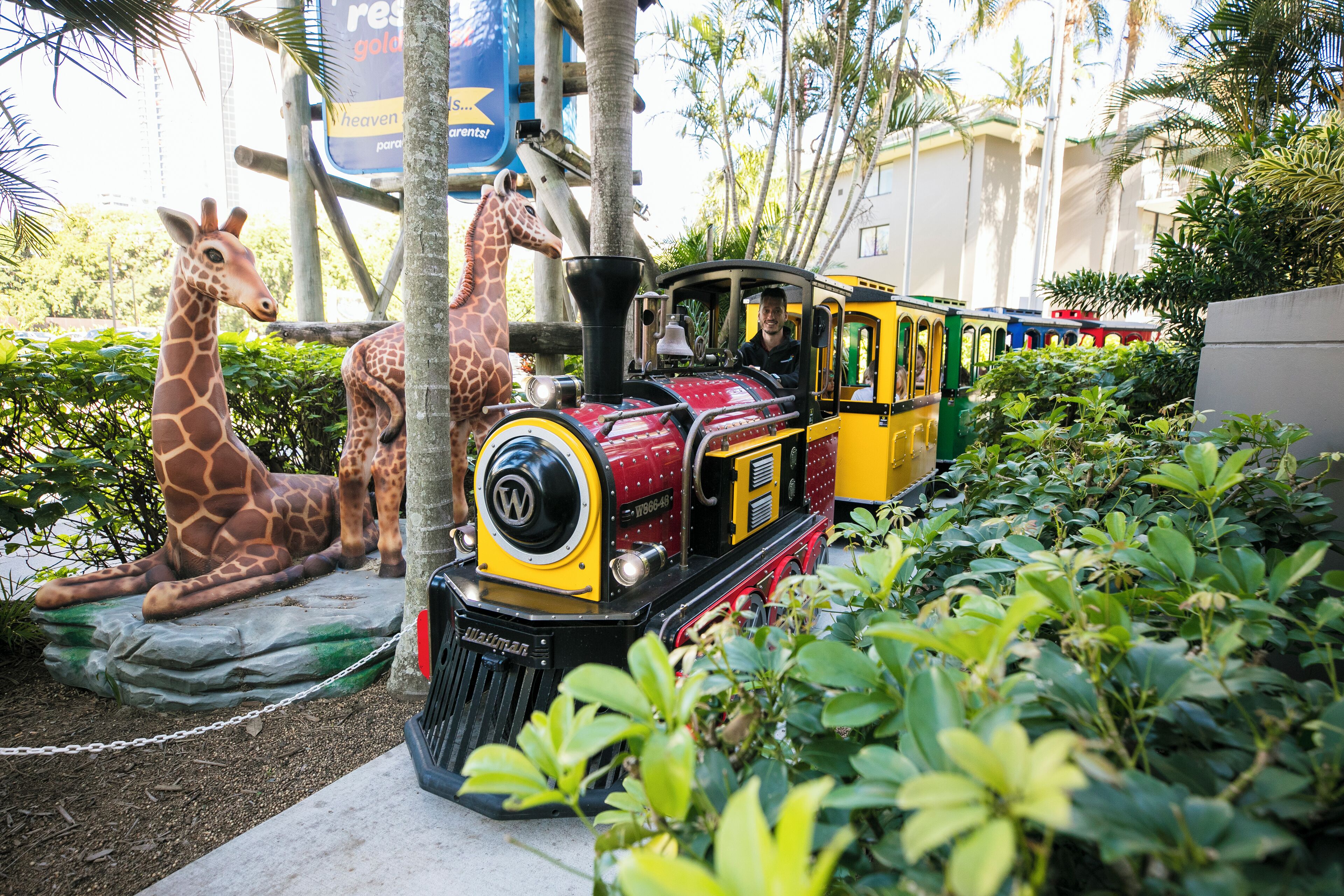 Um trem em miniatura com locomotiva preta e amarela, passando por duas estátuas de girafa.