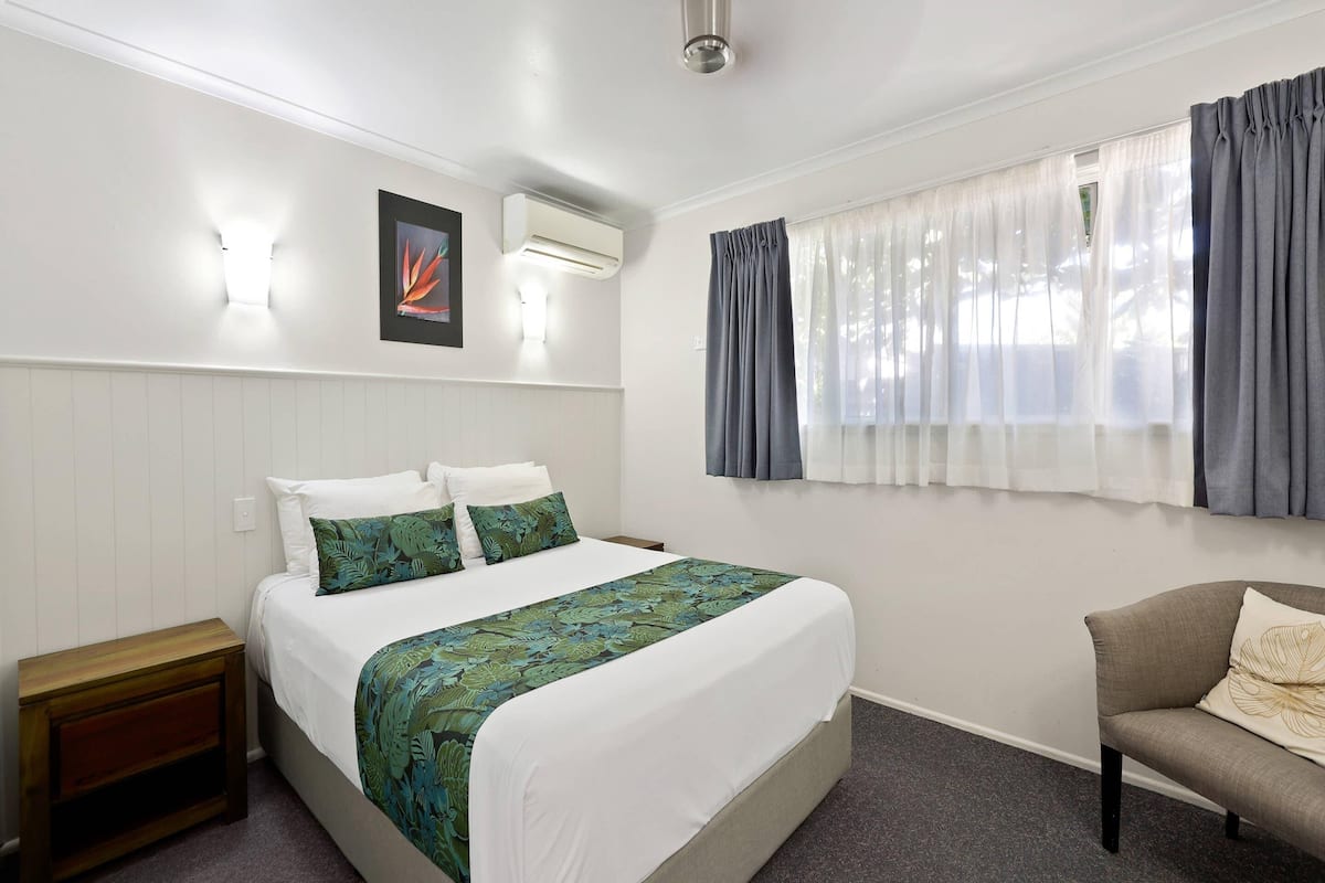 Quarto de hotel com cama, mesa de cabeceira, cadeira, janela com cortinas e iluminação na parede.
