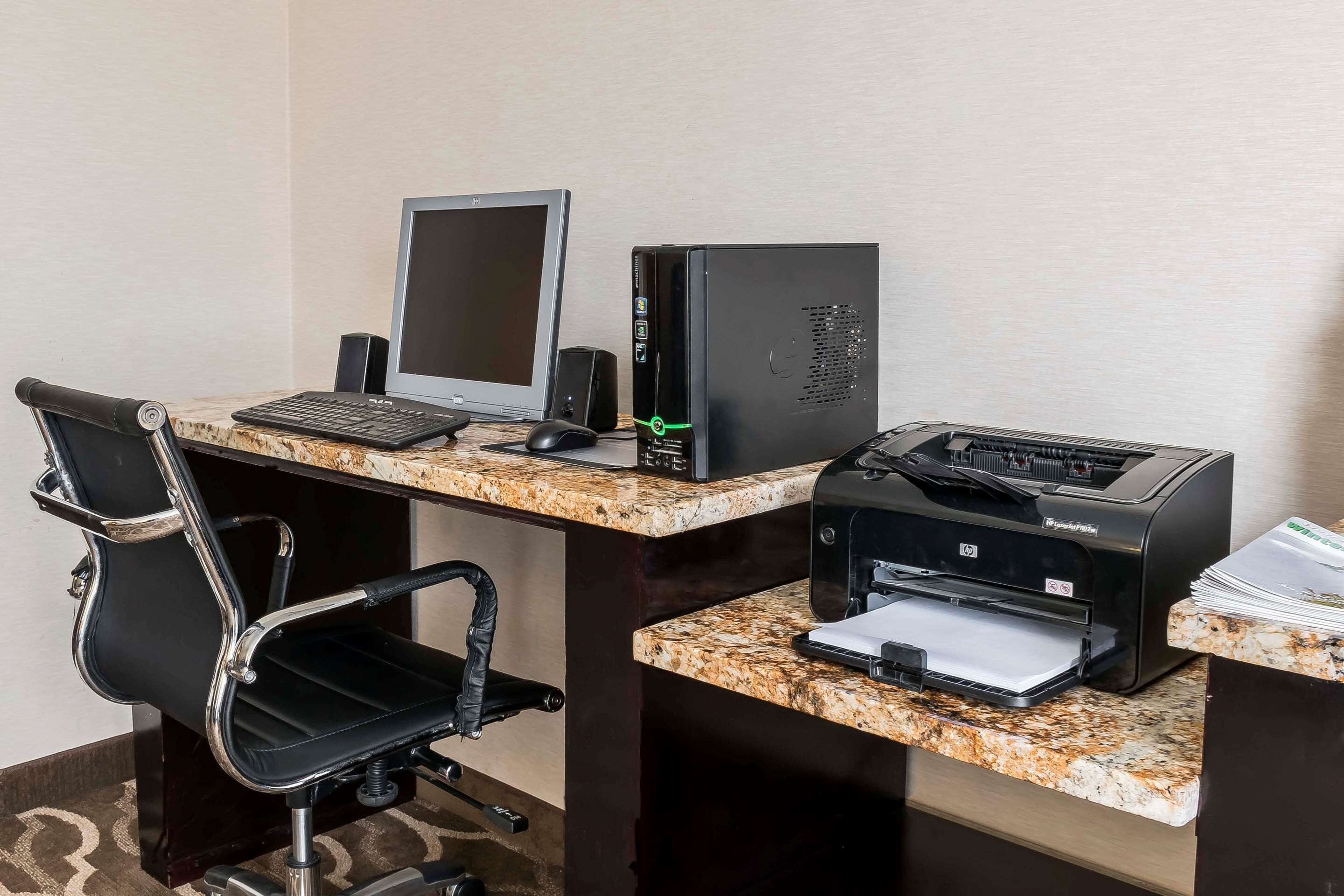 Um espaço de trabalho em hotel, com computador de mesa, monitor, impressora e uma cadeira.