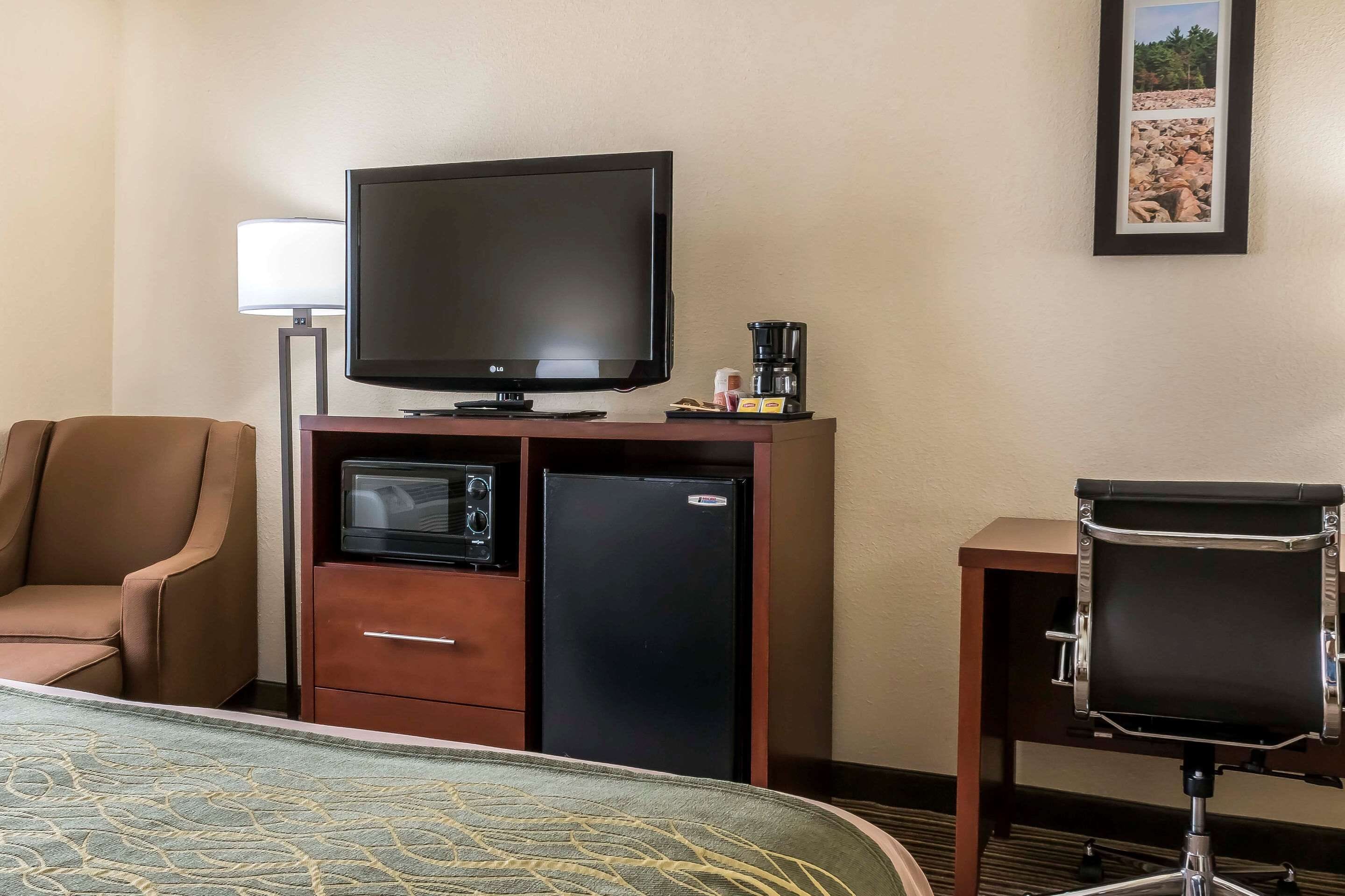 Quarto de hotel com cama, televisão, frigorífico, máquina de café, poltrona e secretária.