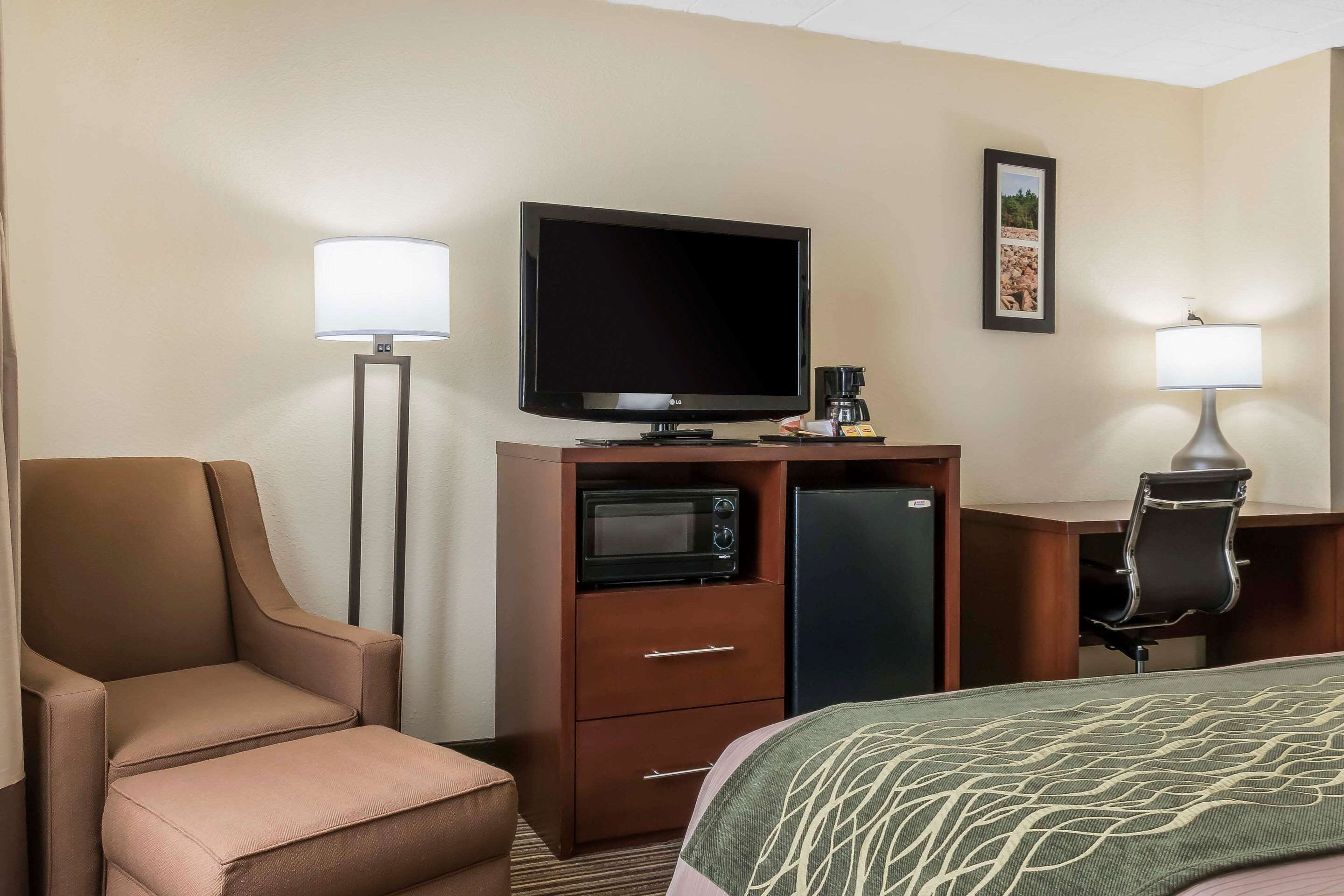 Quarto de hotel com cama, escrivaninha com computador, televisão, micro-ondas, frigorífico e duas luminárias.