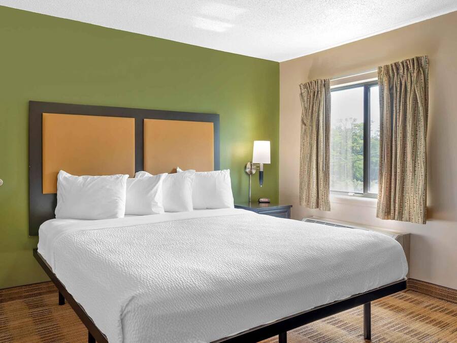 Quarto de hotel com uma cama grande, mesa de cabeceira com abajur, janela com cortinas e parede verde.