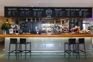 Uma área de bar com um quadro de menu listando bebidas como Bacardi Mojito, Pina Colada e Long Island Ice Tea. Há banquetas de bar e uma variedade de garrafas e copos nas prateleiras.