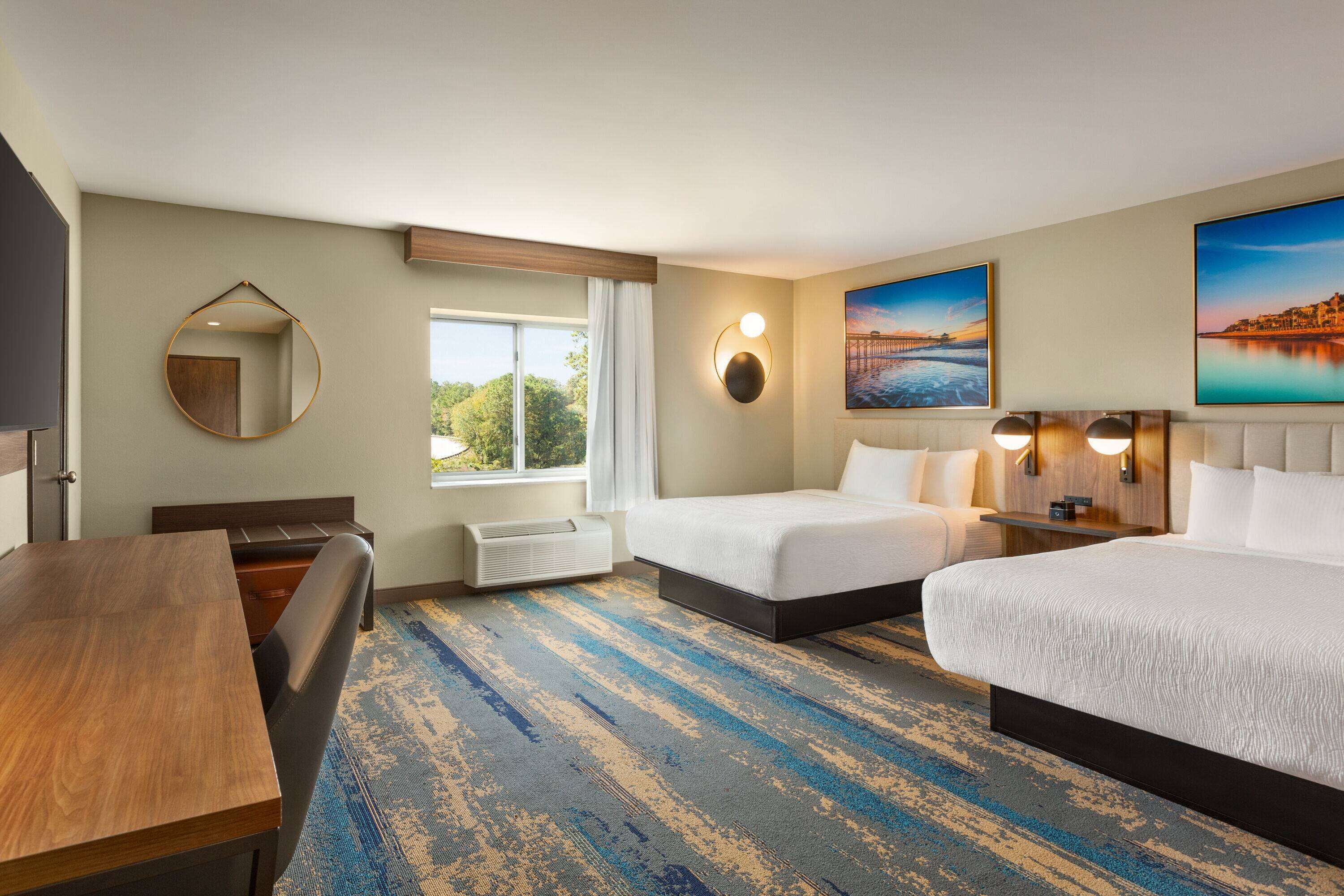 Quarto de hotel com duas camas, uma escrivaninha, uma cadeira, uma janela grande e vista para o exterior.