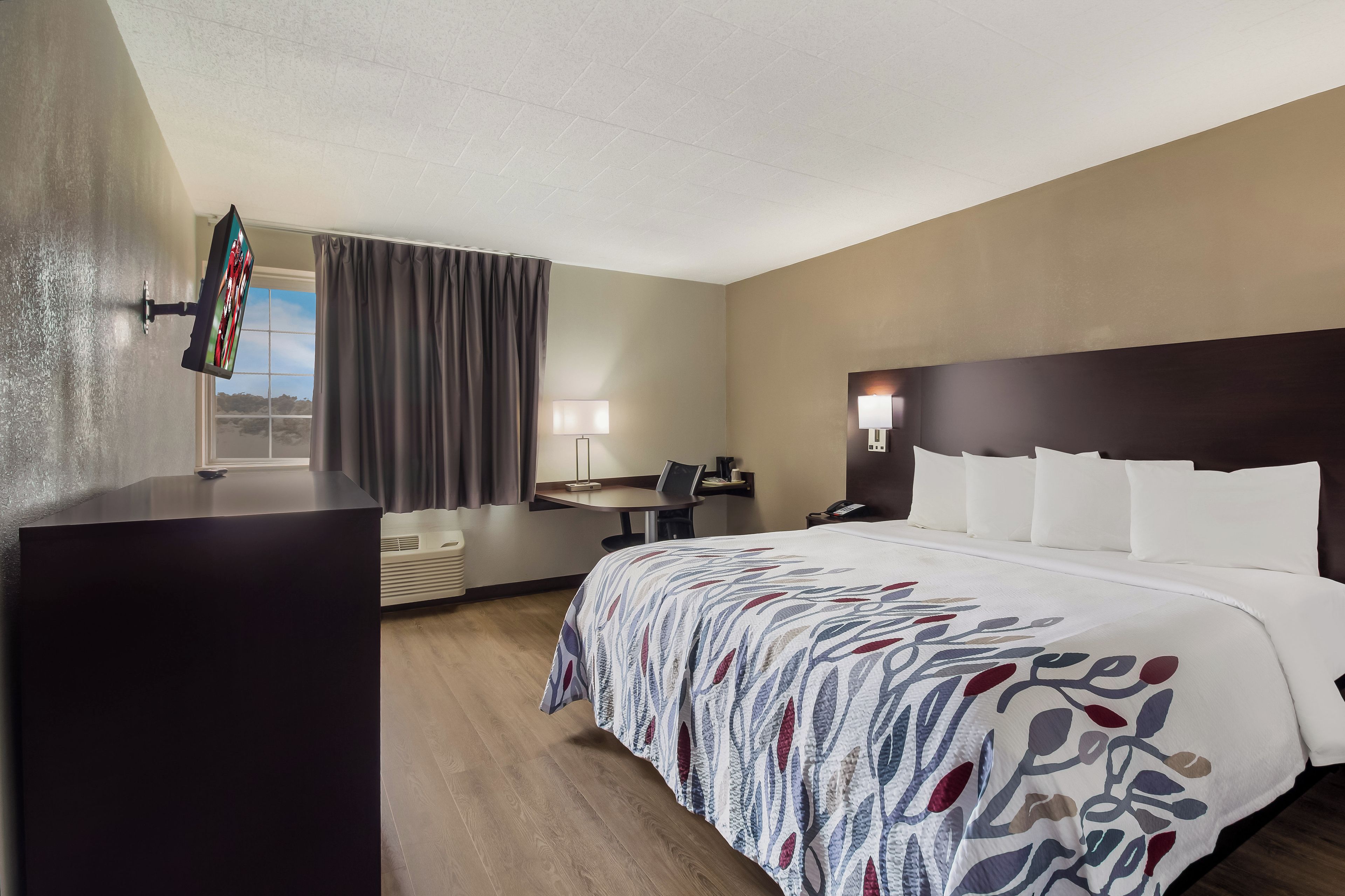 Quarto de hotel com uma cama grande, uma mesa de escritório com um laptop, uma TV fixada na parede e uma janela com cortinas.