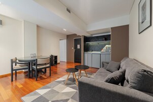 Uma sala de estar moderna com um sofá cinzento, uma mesa de jantar com cadeiras e uma área de cozinha com armários e bancada.