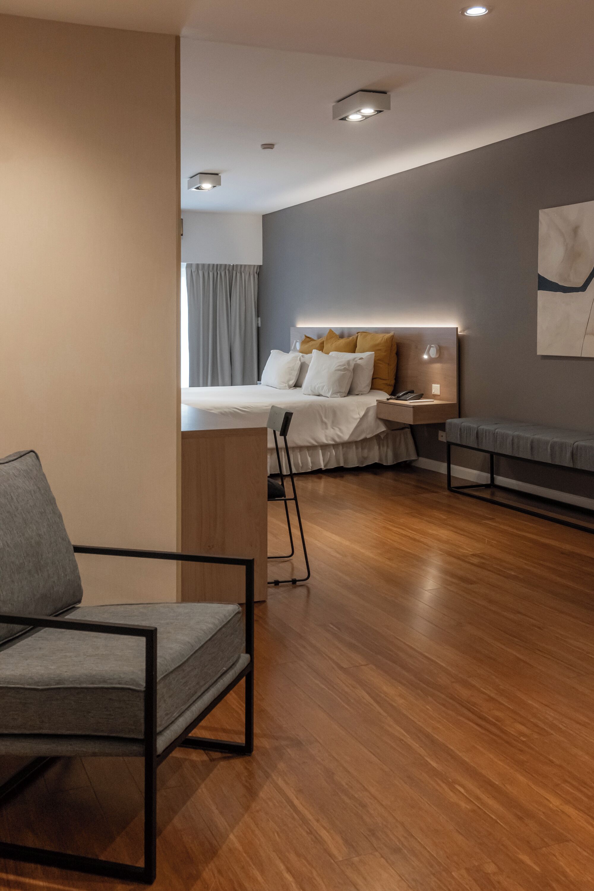 Quarto de hotel moderno com piso de madeira, cama com roupa de cama branca e almofadas amarelas, mesa de cabeceira com abajur e poltrona cinza.