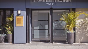 Exterior do Hotel Savoy Double Bay, com um grande logotipo em forma de 'S' nas portas de vidro, uma placa emoldurada na parede e vasos de plantas na frente.