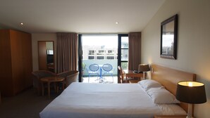 Quarto de hotel com uma cama grande, uma mesa, uma cadeira e uma varanda com vista para edifícios.