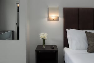 Quarto de hotel com cama, mesa de cabeceira com um vaso de flores, controle remoto e luminária na parede.