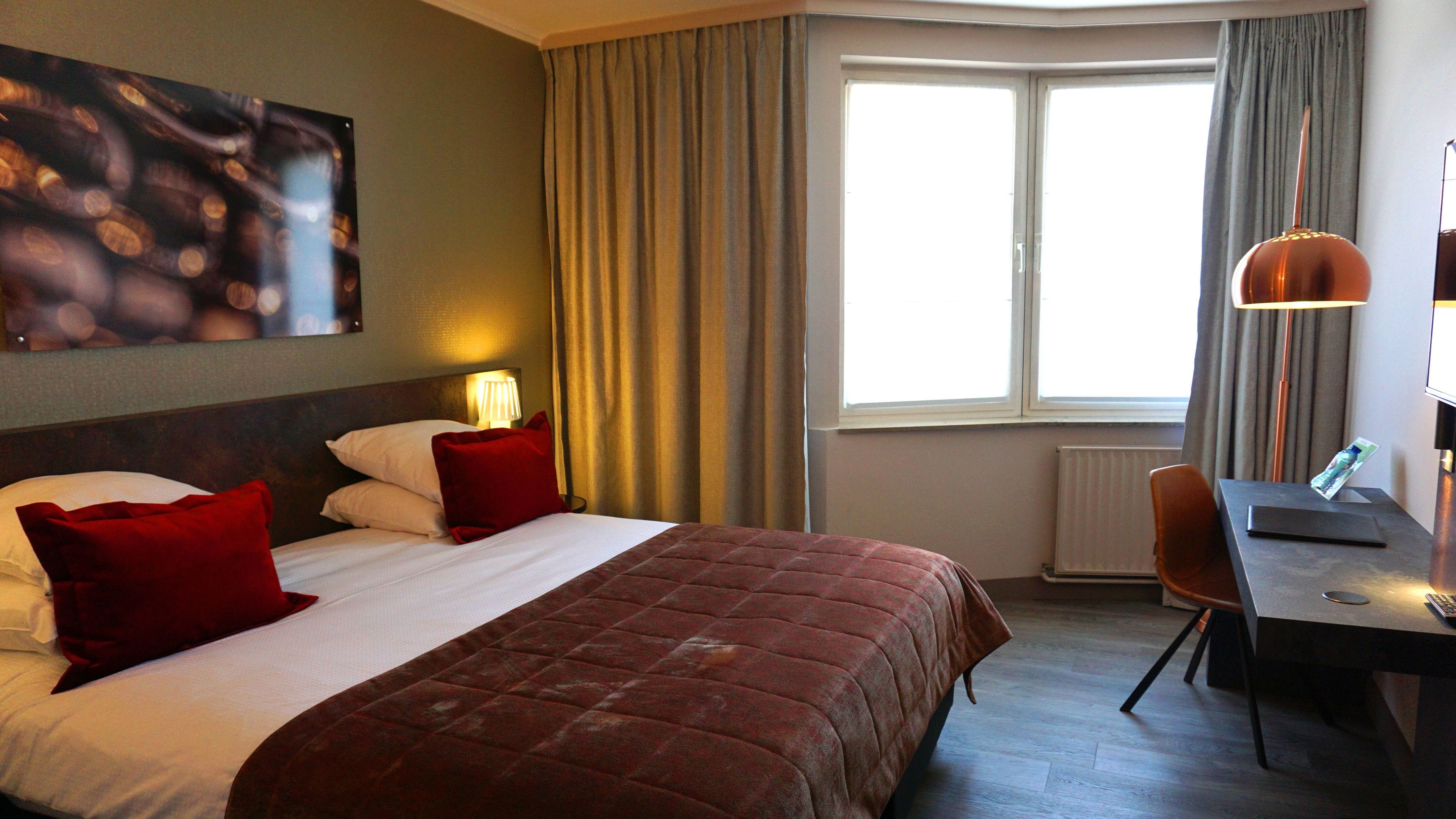 Quarto de hotel com uma cama grande, duas almofadas vermelhas, uma escrivaninha com luminária e uma janela com cortinas.