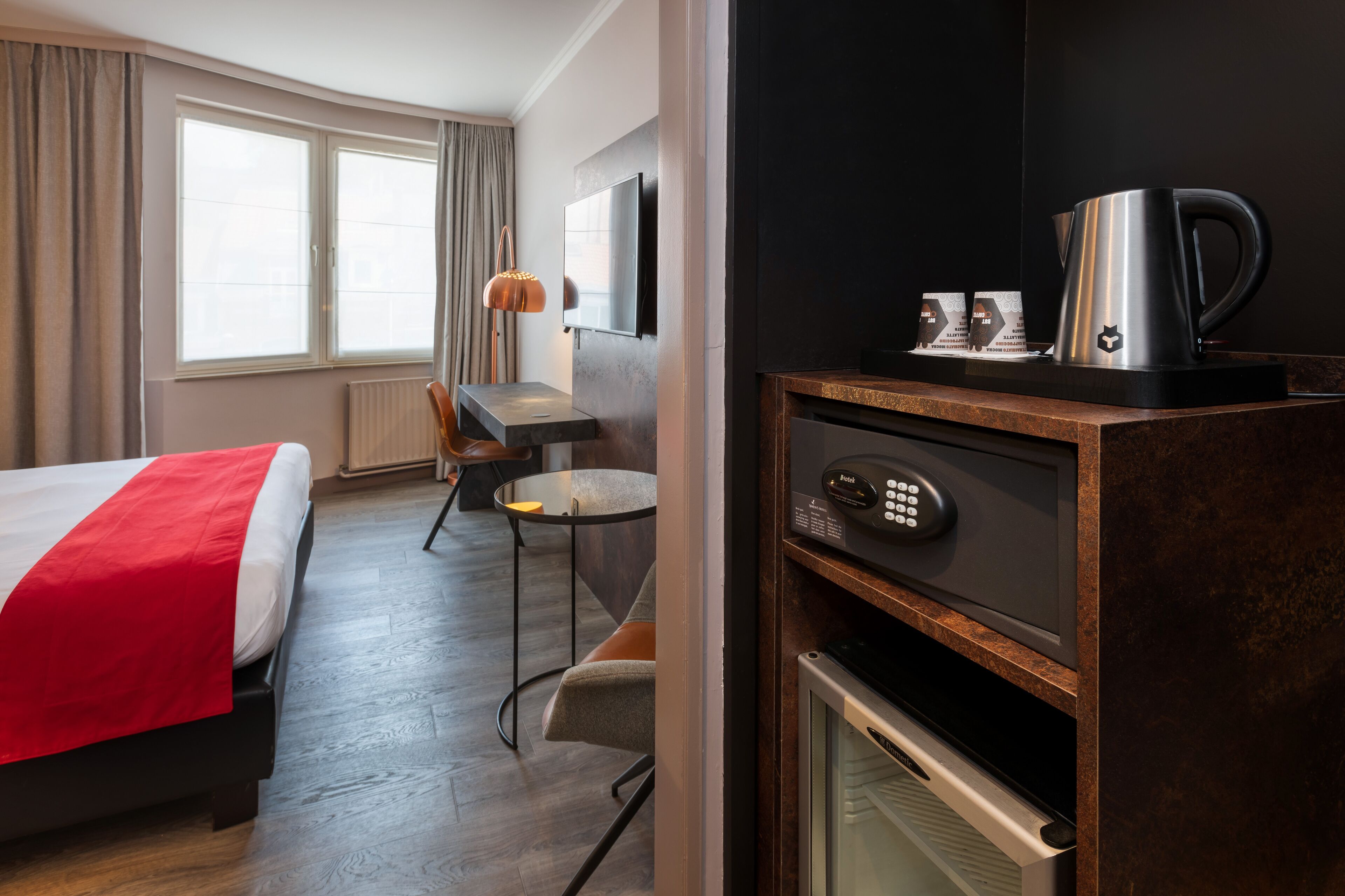 Quarto de hotel com cama, escrivaninha, cadeira, cafeteira e televisão.