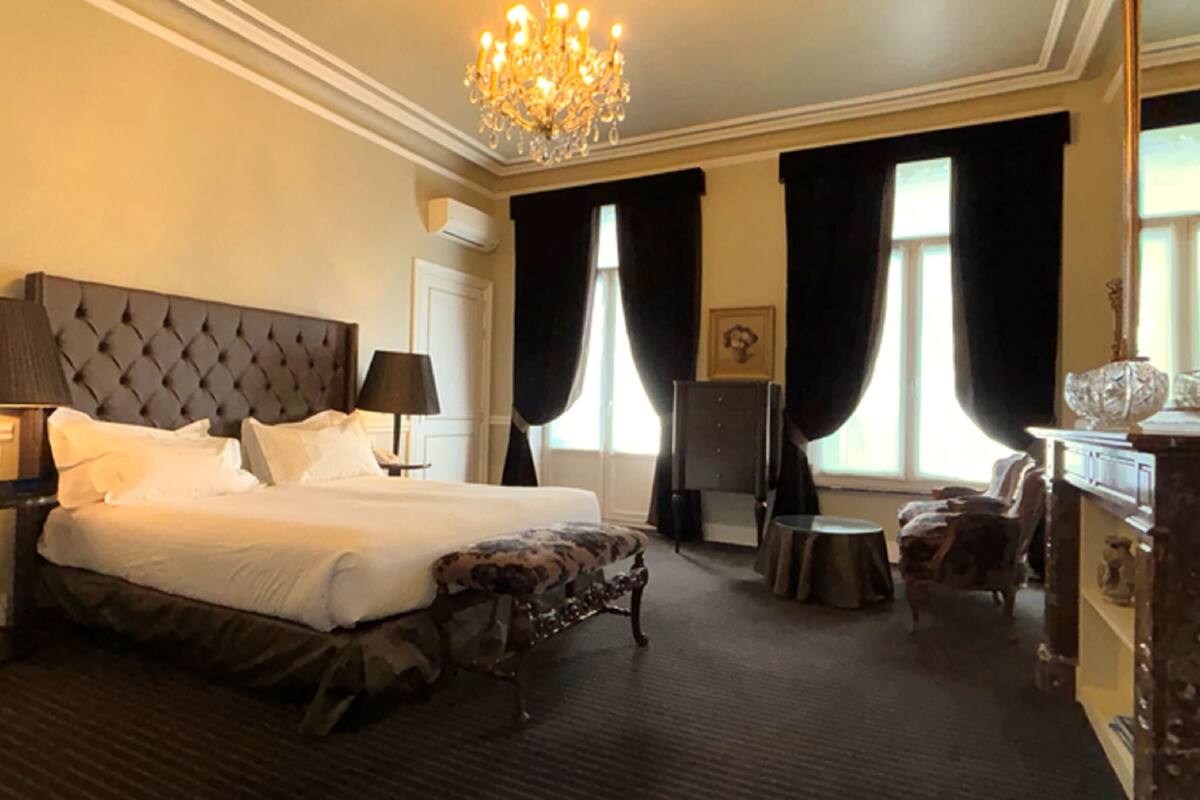 Quarto de hotel com uma cama grande, duas luminárias de cabeceira, um lustre, uma mesinha redonda, uma cadeira e uma cômoda.