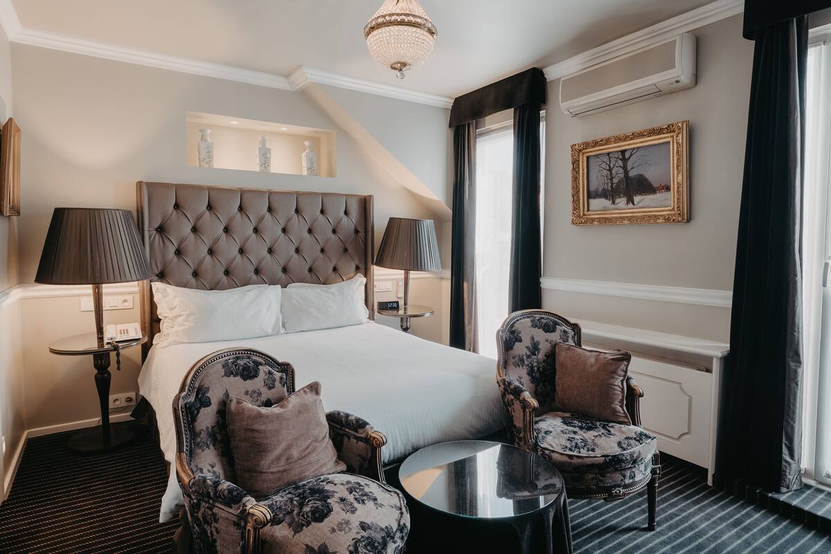 Quarto de hotel com uma cama grande, duas poltronas estofadas, uma mesa redonda pequena, um abajur e um quadro na parede.