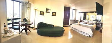 Quarto de hotel com uma cama, uma poltrona verde, uma mesinha com um vaso de flores e uma área de refeições visível pela janela.