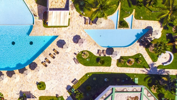 Vista aérea de um resort com uma grande piscina, espreguiçadeiras, guarda-sóis e palmeiras.
