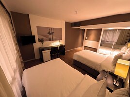 Quarto de hotel com cama, escrivaninha com cadeira, mini-frigorífico, televisão fixada na parede e um espelho grande.