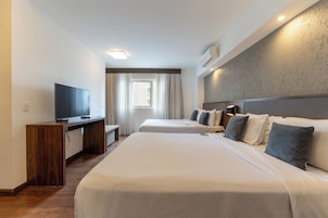 Quarto de hotel com duas camas, uma mesa de trabalho com televisão de tela plana, uma janela com cortinas e um ar condicionado na parede.