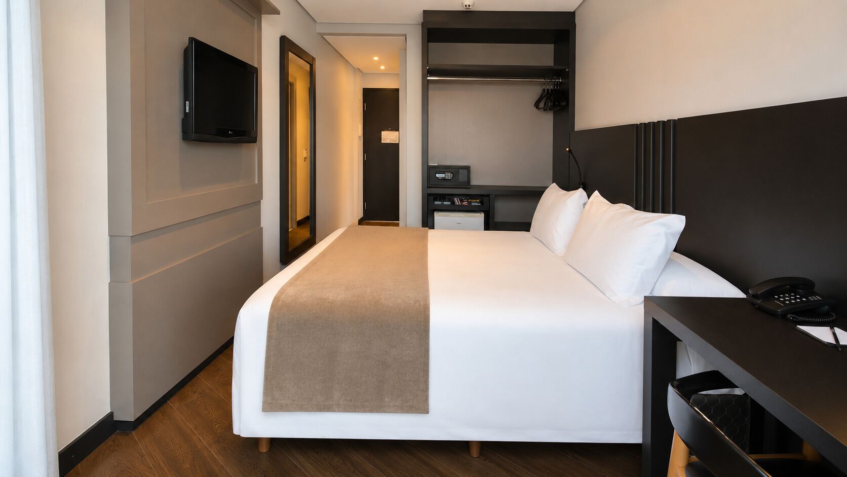 Quarto de hotel com cama, mesa de cabeceira, televisão e escrivaninha com telefone.