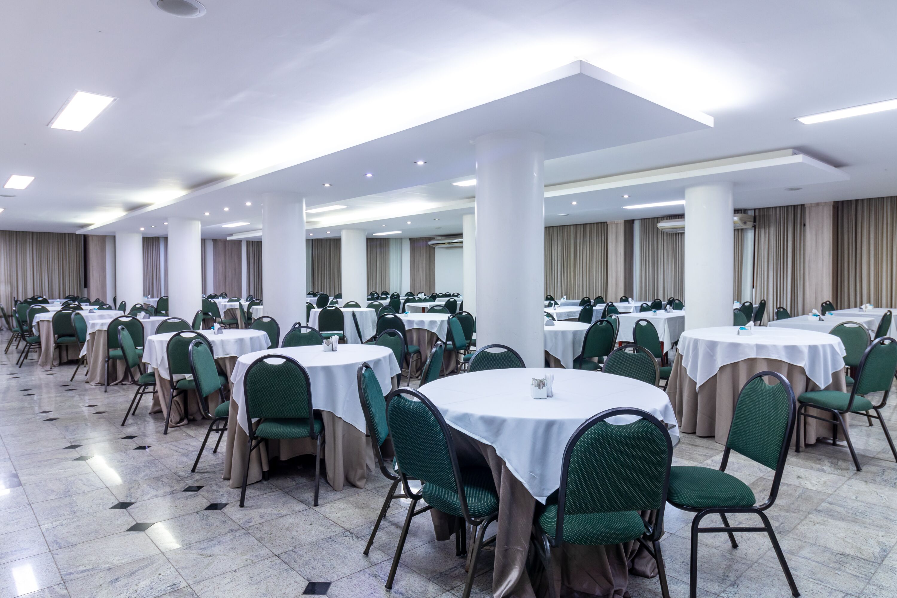 Um amplo salão de festas com mesas redondas e cadeiras verdes, toalhas de mesa brancas e cortinas bege.