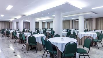 Um amplo salão de festas com mesas redondas e cadeiras verdes, toalhas de mesa brancas e cortinas bege.