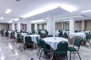 Um amplo salão de festas com mesas redondas e cadeiras verdes, toalhas de mesa brancas e cortinas bege.