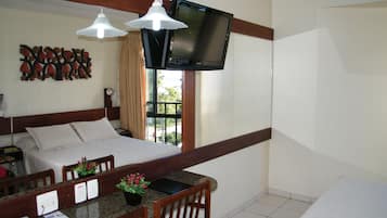 Quarto de hotel com cama, escrivaninha com telefone e planta, televisão na parede e janela com cortinas.