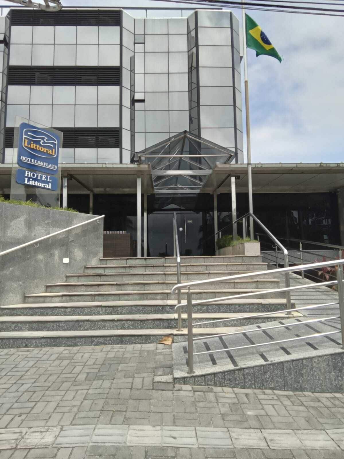 Entrada de hotel com placa da Litoral Hotels & Flats, uma escadaria e a bandeira do Brasil.