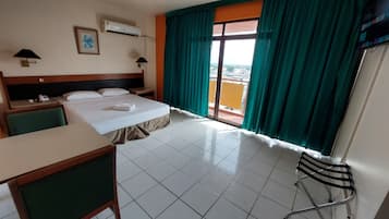 Quarto de hotel com cama, escrivaninha, cadeira, televisão e varanda com cortinas.