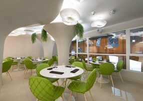 Restaurante moderno com mesas redondas e cadeiras verdes, janelas amplas e plantas pendentes.