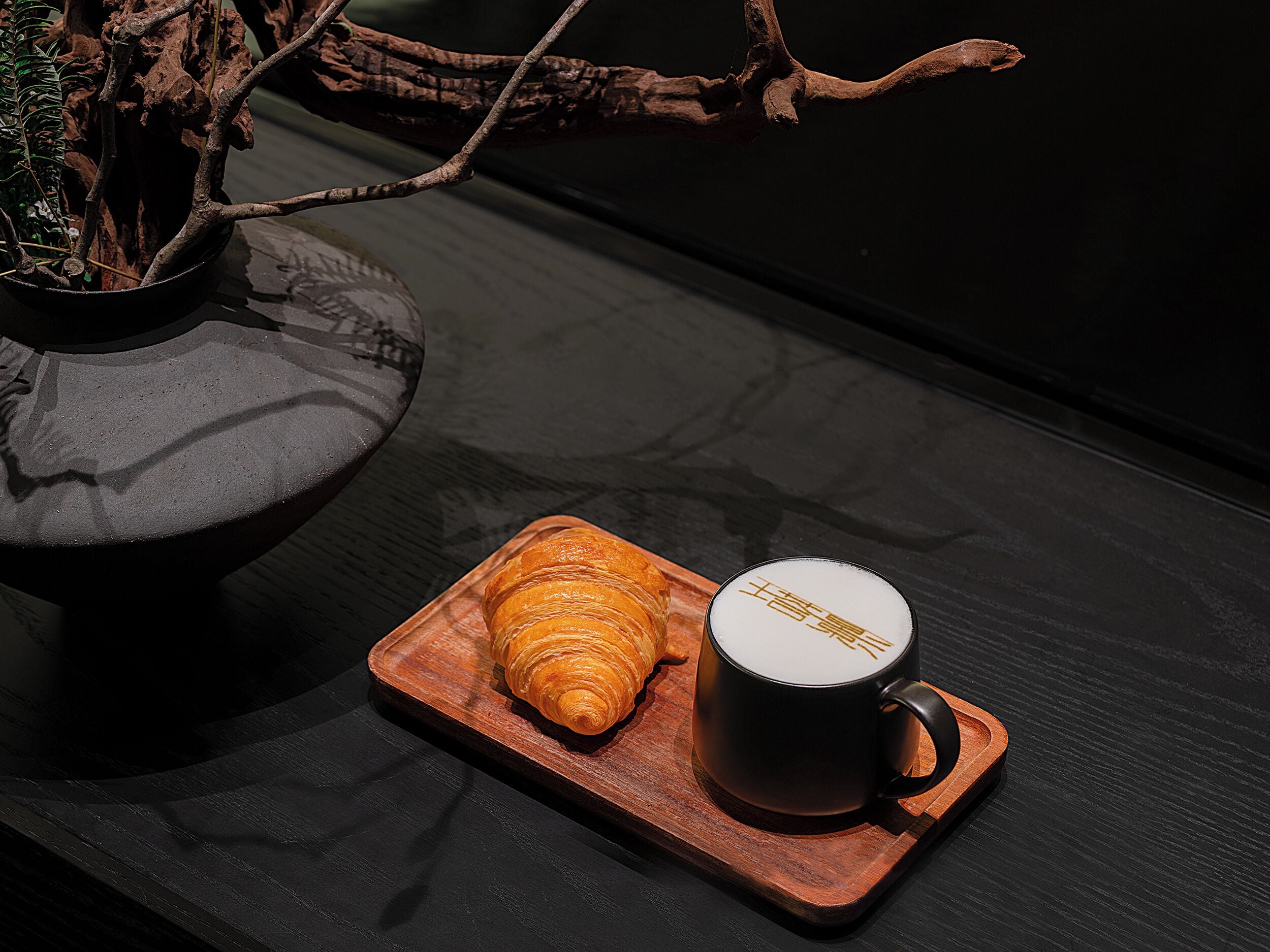 Um croissant e uma chávena numa bandeja de madeira sobre uma superfície escura.