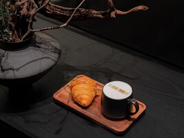 Um croissant e uma chávena numa bandeja de madeira sobre uma superfície escura.