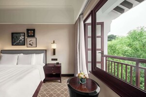 Quarto de hotel com cama, mesa de cabeceira, luminária e janela com vista para vegetação.