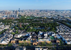 Vista aérea de um horizonte urbano com intenso desenvolvimento, uma combinação de edifícios altos e baixos, e uma área verde central com arquitetura tradicional.
