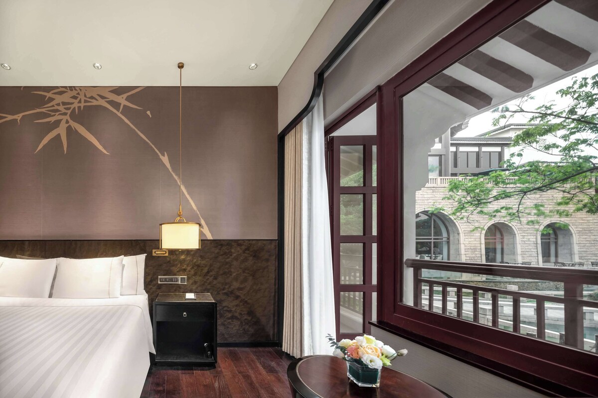 Quarto de hotel com uma grande janela, uma cama, uma mesa de cabeceira, um abajur e um vaso com flores.