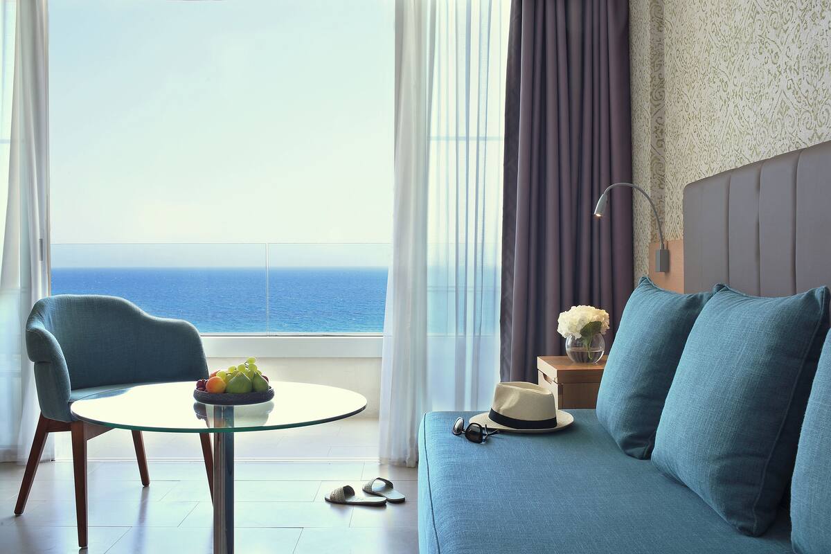 Quarto com vista para o mar, um sofá, uma poltrona, uma mesa com frutas e um chapéu no sofá.