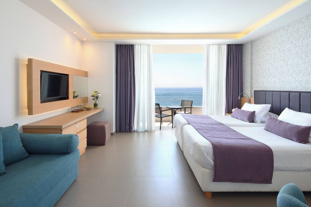 Quarto de hotel moderno com uma cama grande, um sofá, uma TV e vista para o mar.