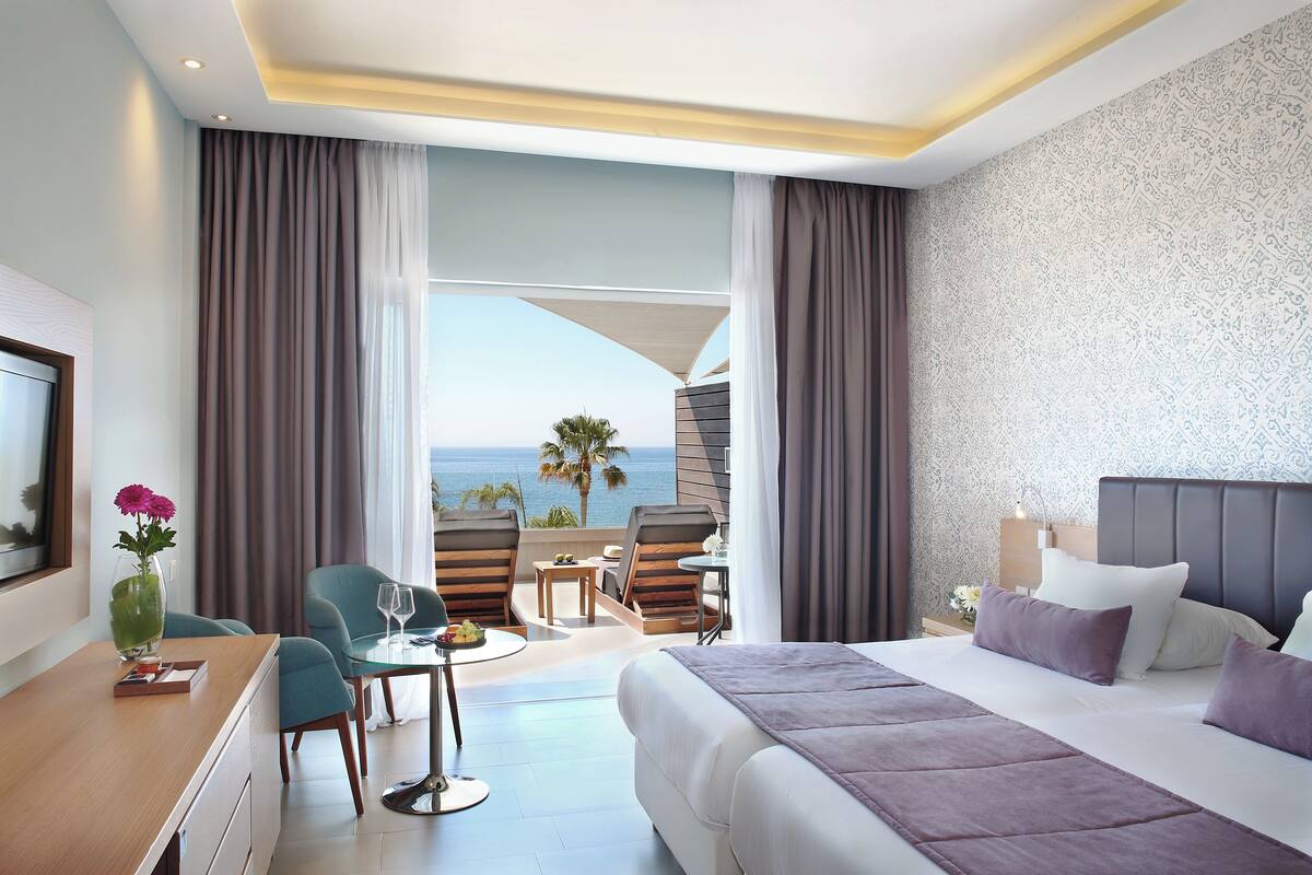 Quarto de hotel com uma cama, uma escrivaninha, uma cadeira, uma mesinha e vista para o mar.