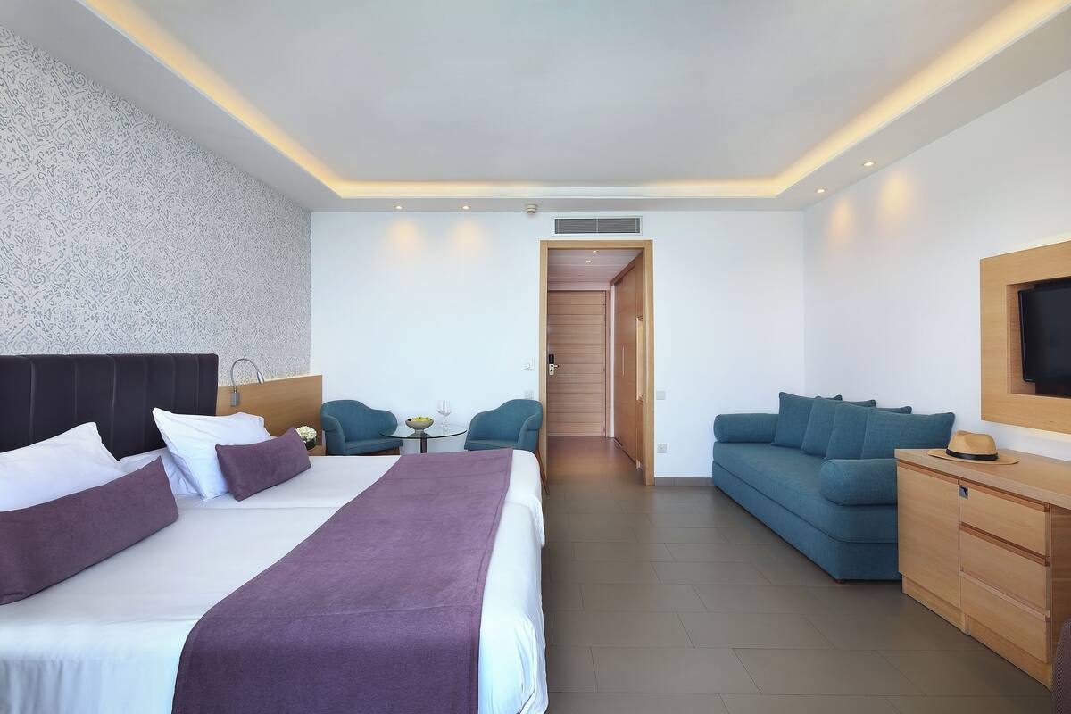 Quarto de hotel moderno com uma cama grande, um sofá azul, um armário de madeira e uma televisão na parede.