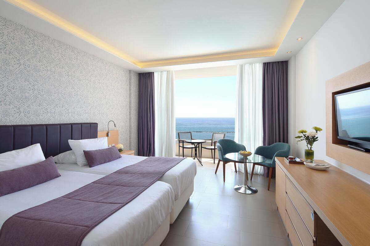 Quarto de hotel com duas camas, varanda com vista para o mar, escrivaninha com televisão e uma pequena mesa com cadeiras.
