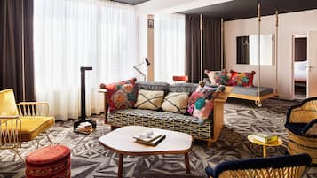 Quarto de hotel moderno com sofás, mesa de centro redonda, um banquinho vermelho e uma cadeira de vime.