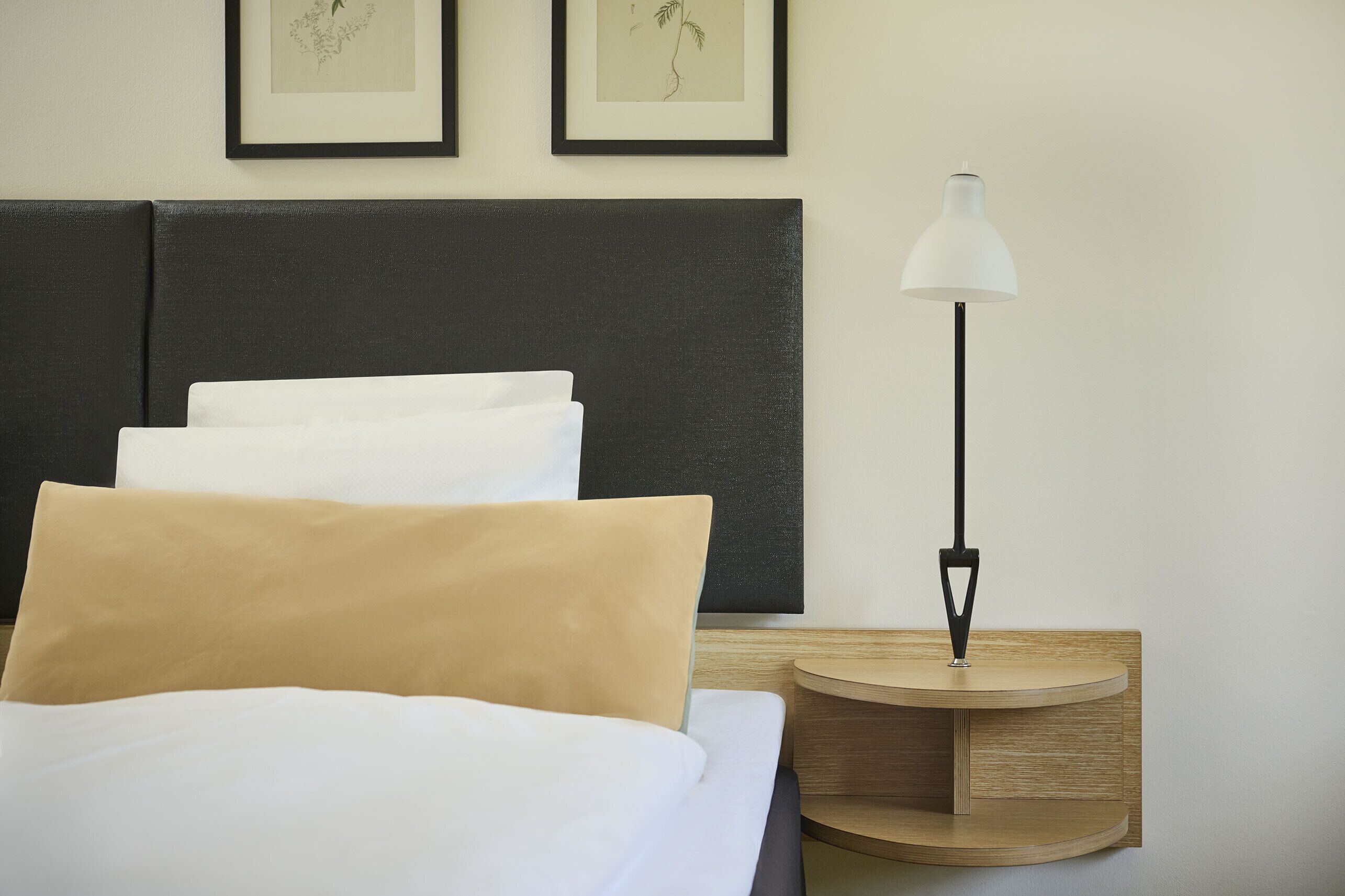 Uma cama bem arrumada com roupa de cama branca e bege, cabeceira escura e uma mesa de cabeceira com abajur.