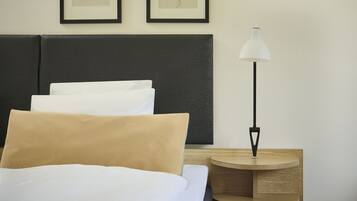 Uma cama bem arrumada com roupa de cama branca e bege, cabeceira escura e uma mesa de cabeceira com abajur.