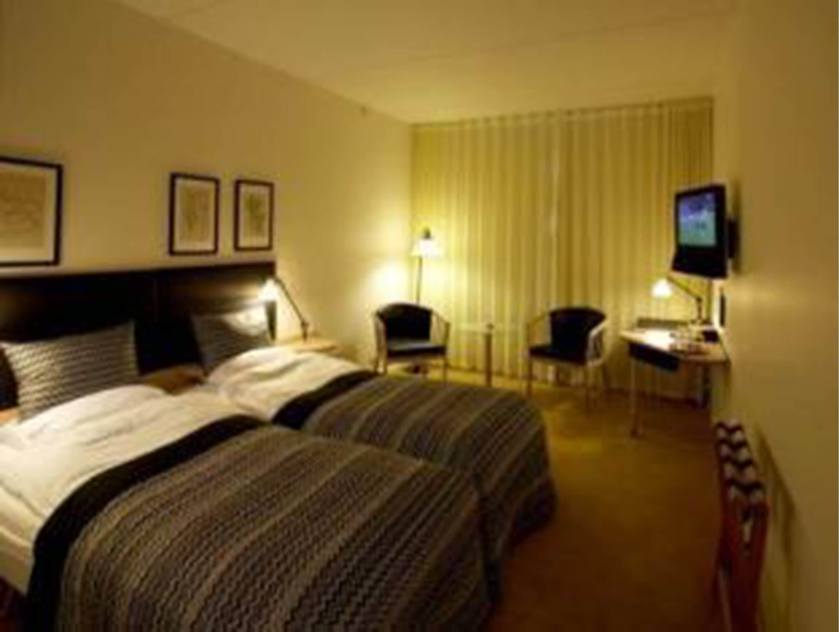 Quarto de hotel com duas camas, uma mesa de trabalho com computador, uma cadeira e televisão.
