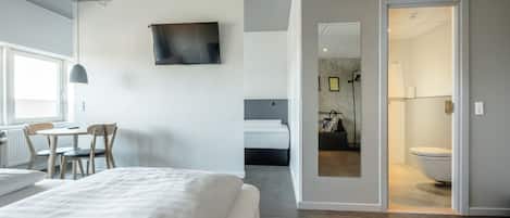 Quarto de hotel moderno com uma cama grande, uma escrivaninha com cadeira, televisão na parede e casa de banho visível por uma porta aberta.