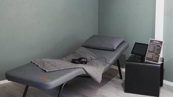 Sala de exames com uma mesa de exame individual, uma pequena mesa lateral preta e um quadro de informações fixado na parede.
