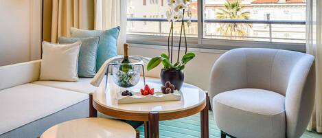 Quarto de hotel moderno com sofá, poltrona e uma pequena mesa com garrafa, planta e frutas.